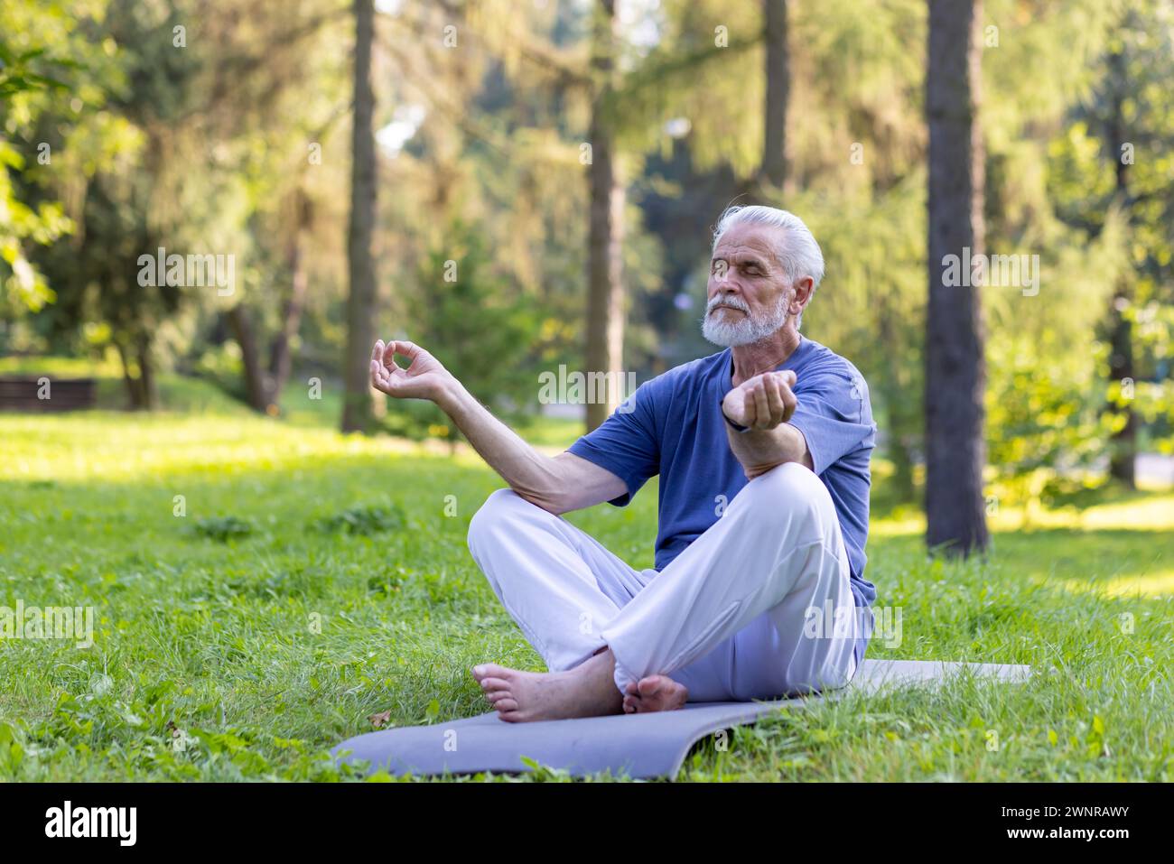 Senior gray-haired man doing yoga in the park, sitting on a mat in the ...