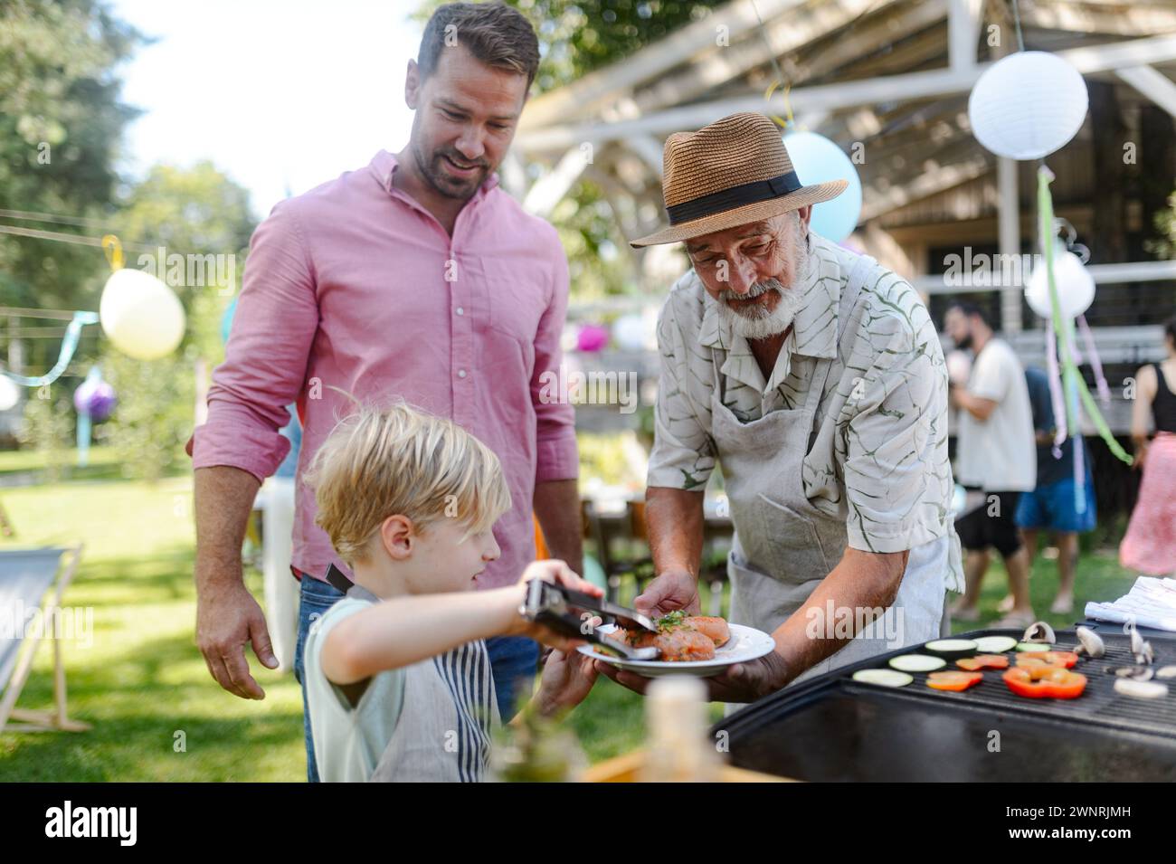 Father, grandfather and son grilling together at garden BBQ. Three generations of men at summer ...