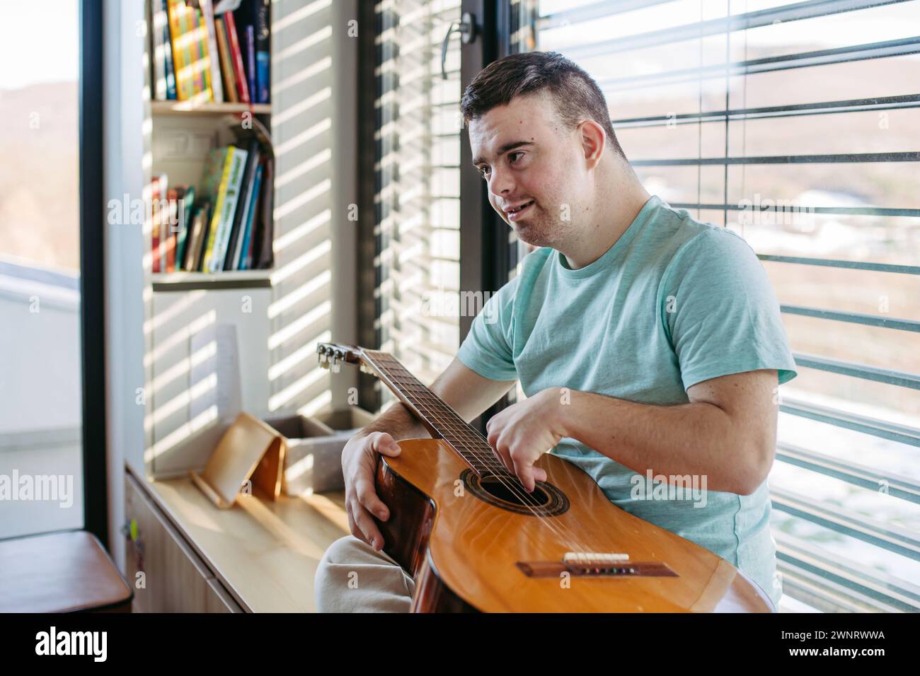 Young man with down syndrome playing acoustic guitar, sitting by window ...