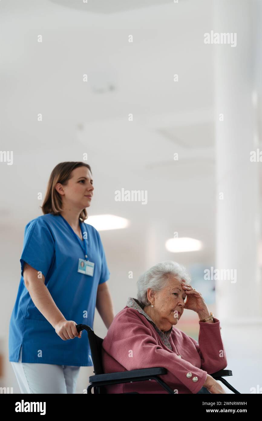 Nurse pushing senior patient in wheelchair across hospital corridor ...