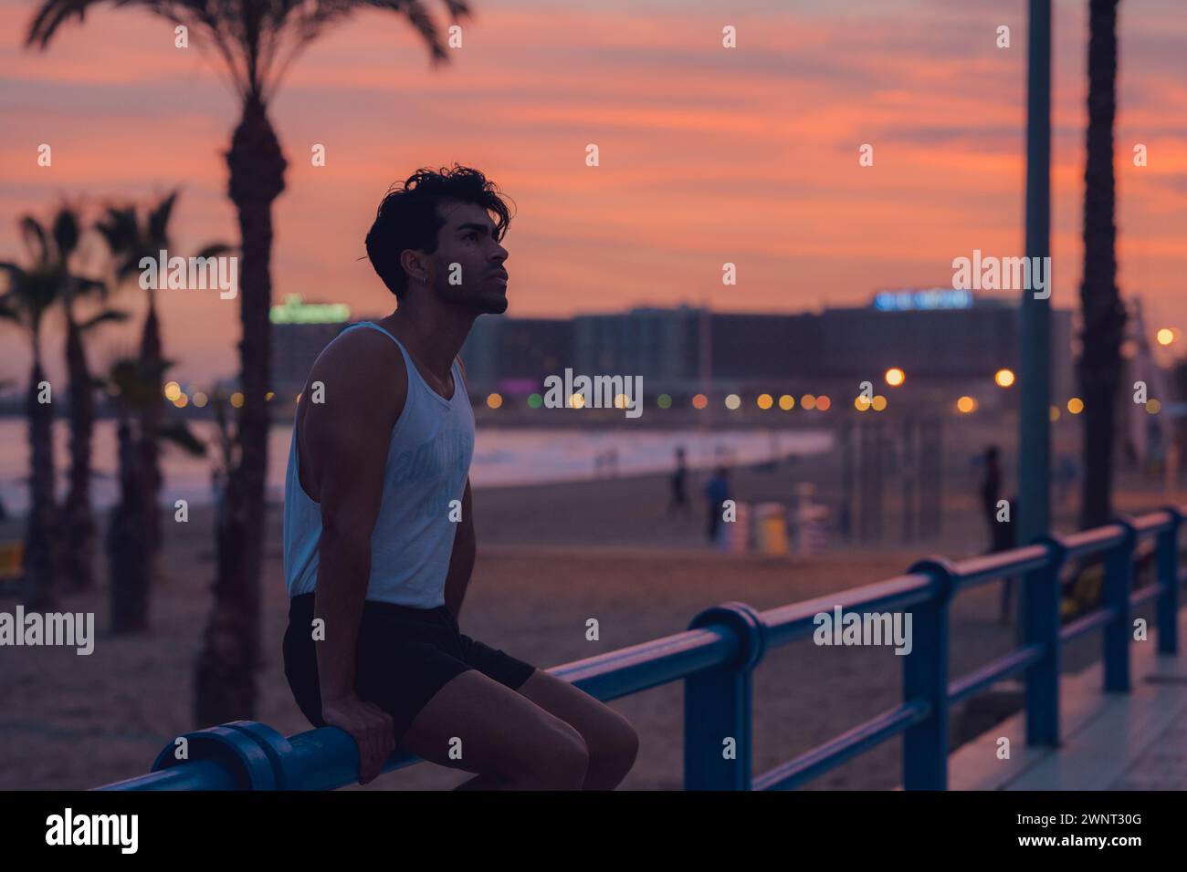 Pensive male athlete resting on a blue railing at beachside at sunset ...
