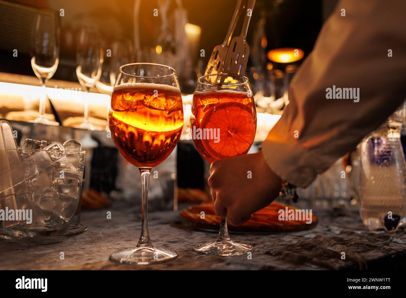 Bartender makes two glasses of cocktail Aperol spritz on bar counter, adds fresh orange slices ...