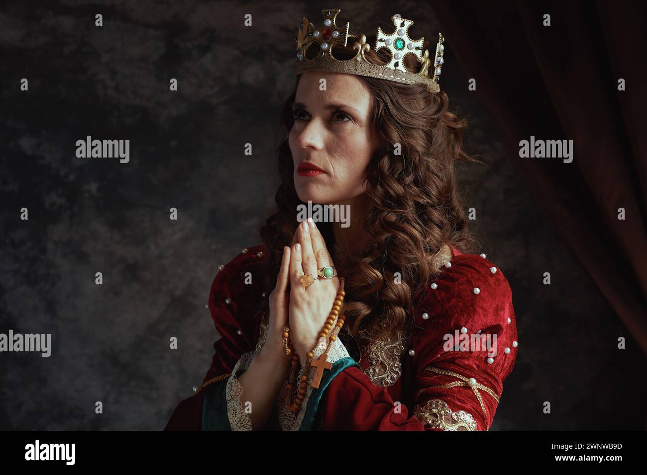 medieval queen in red dress with rosary and crown praying on dark gray ...