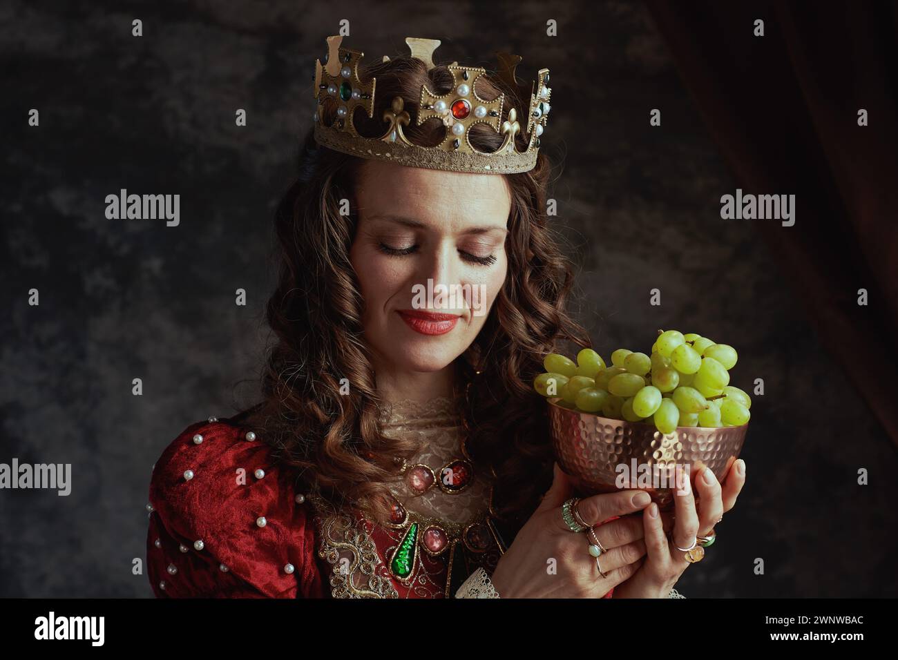 medieval queen in red dress with plate of grapes and crown on dark gray ...