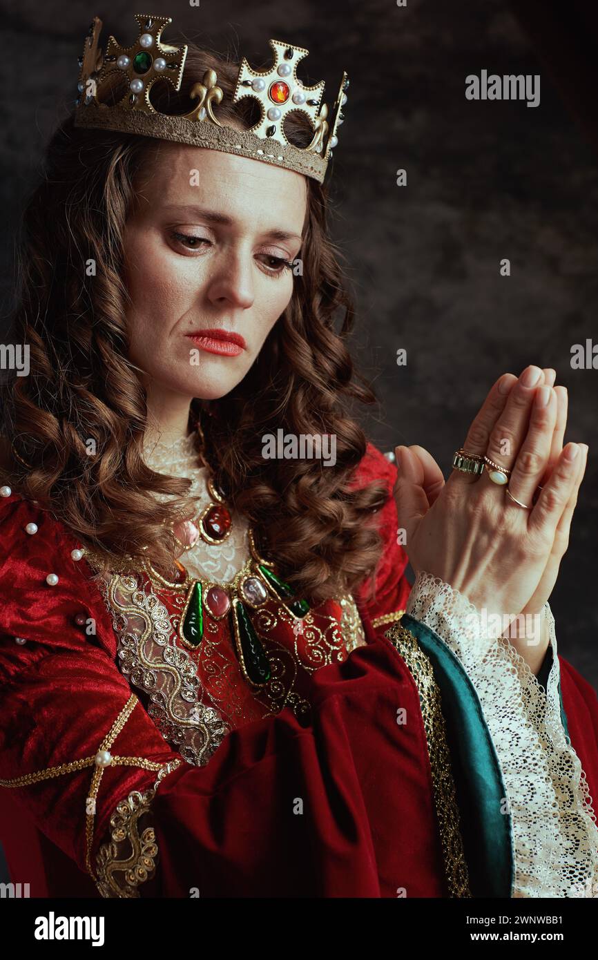 medieval queen in red dress with crown praying on dark gray background ...