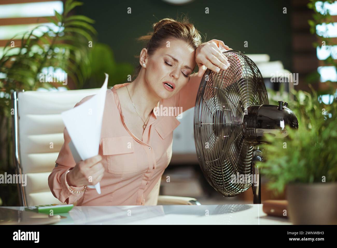 Sustainable workplace. modern middle aged woman employee at work with documents and electric fan ...