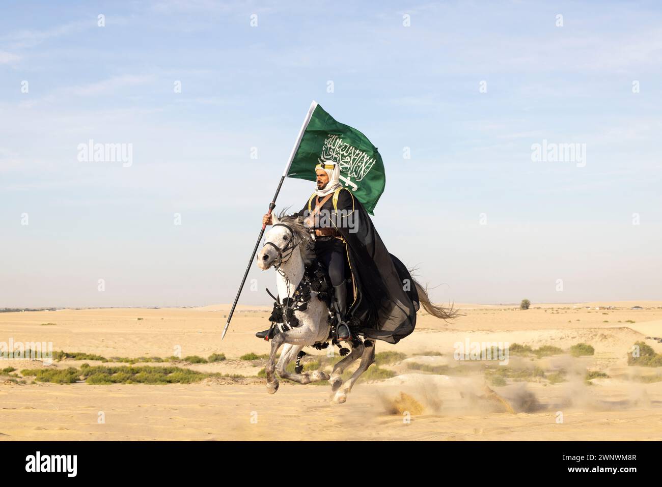 Man in traditional clothing with his horse in a desert, carrying a flag ...