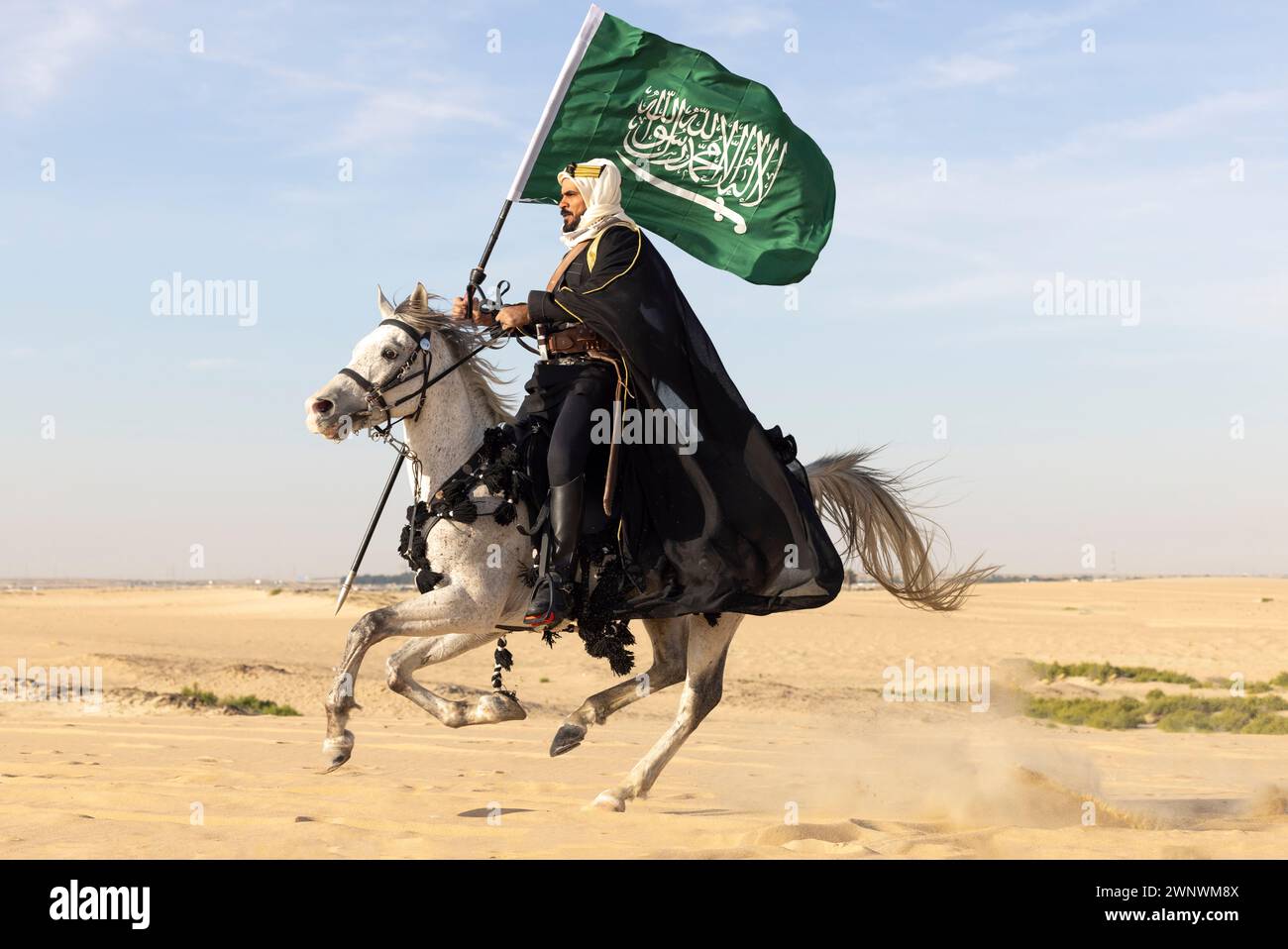 Man in traditional clothing with his horse in a desert, carrying a flag ...