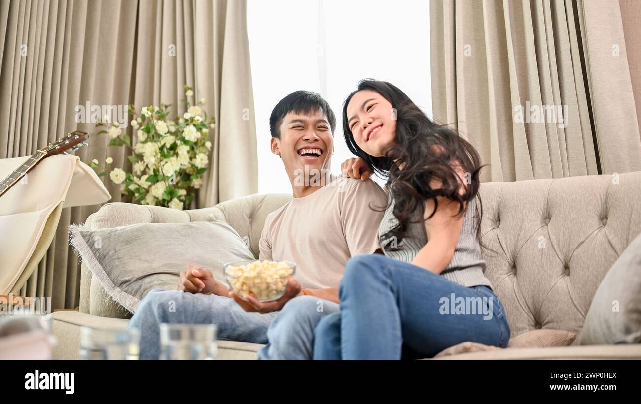 A cheerful and excited young Asian couple is enjoying popcorn while watching an exciting sports ...