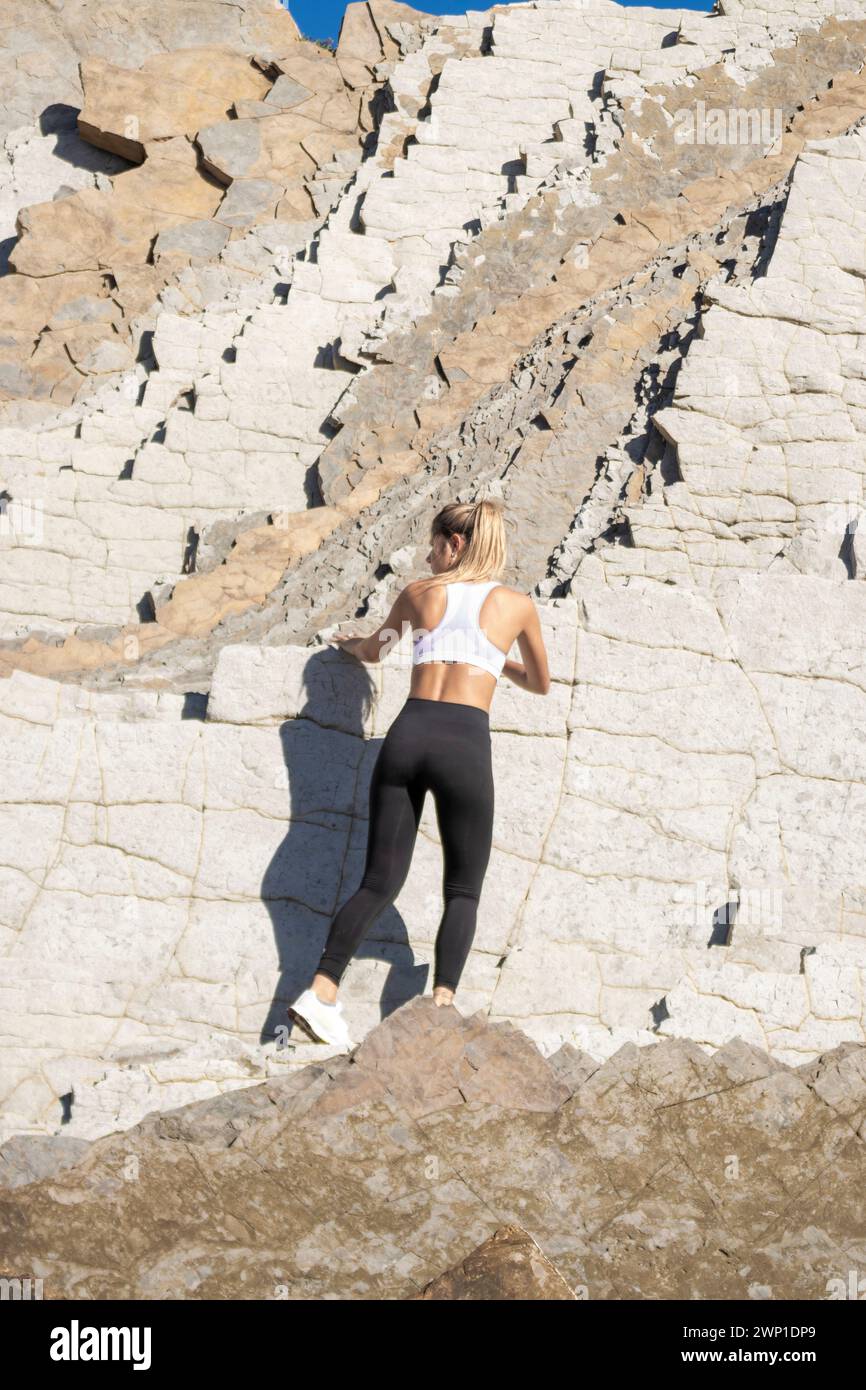 A woman is climbing a rock wall in a white tank top and black pants ...