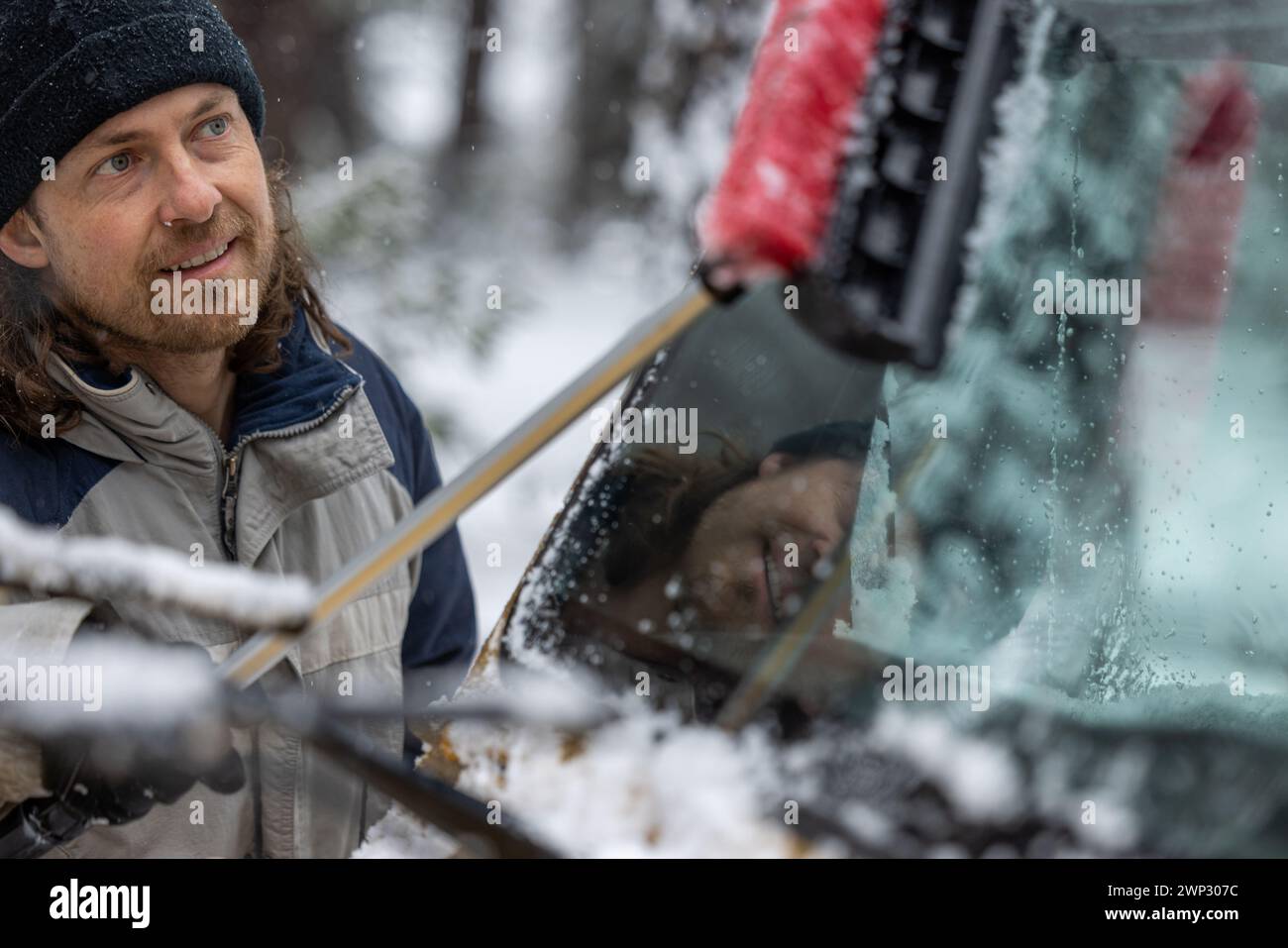 A man is cleaning the windshield of a car with a snow brush. He is ...