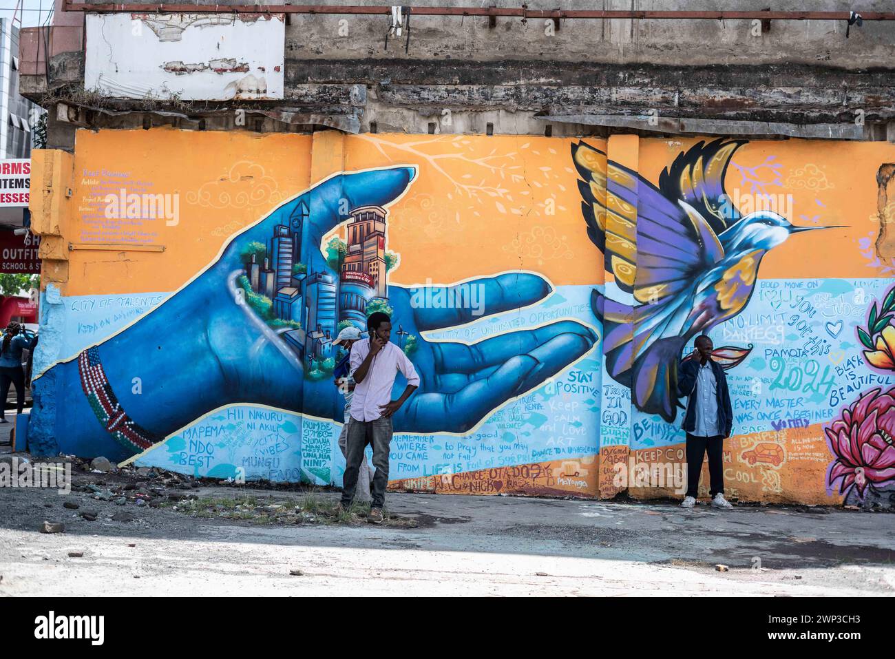 Two men stand in front of murals depicting a hand holding a city and a ...