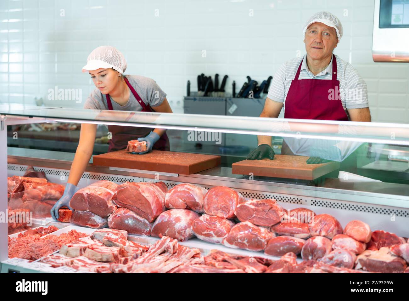 In butcher shop, elderly male vendor in uniform apron, gloves, cap ...