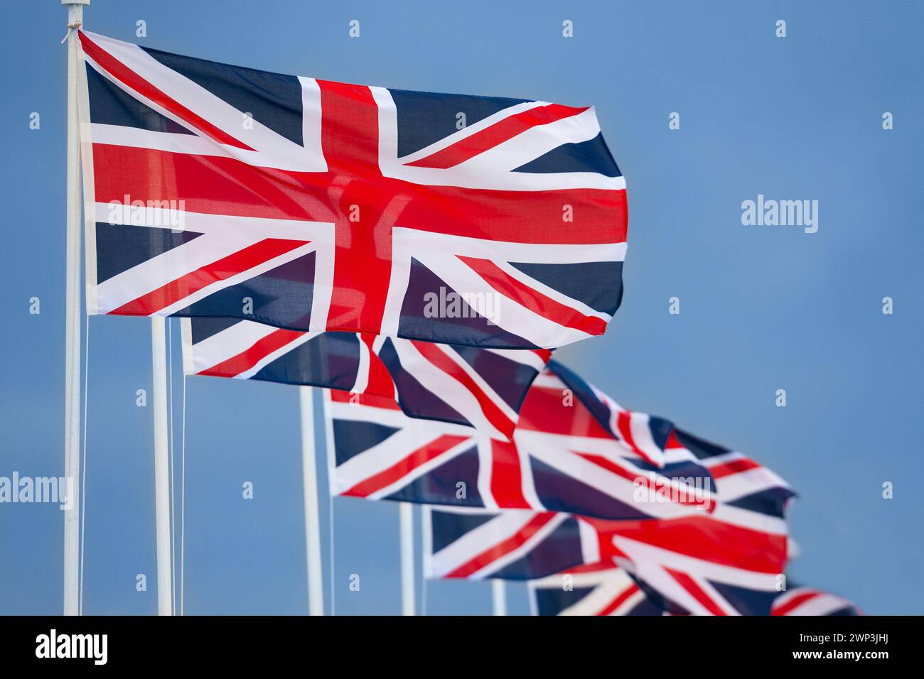 Row of Union Jack British flags flying on flagpoles against a blue sky ...