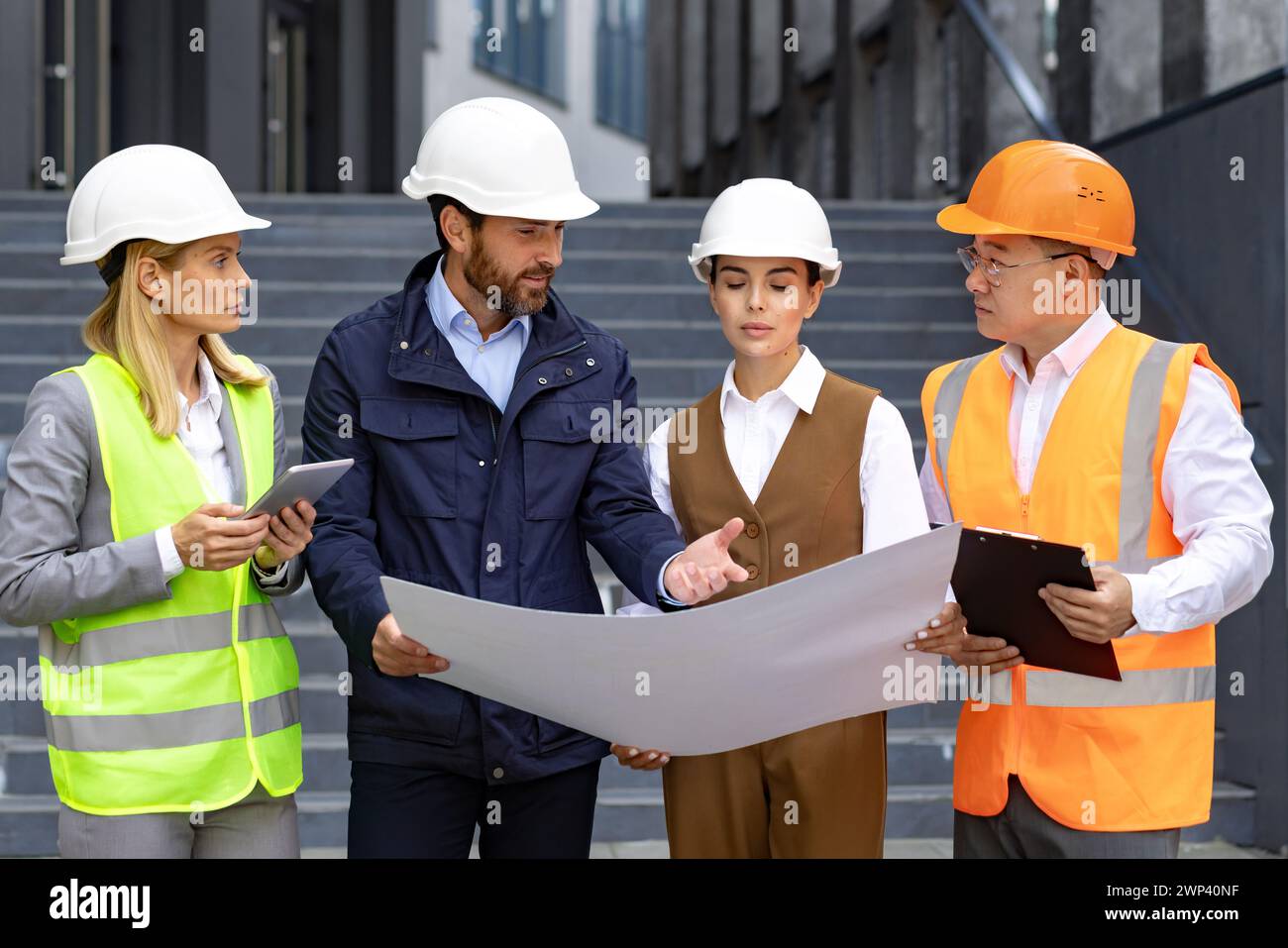 Focused diverse group of engineers with hard hats discussing blueprints ...