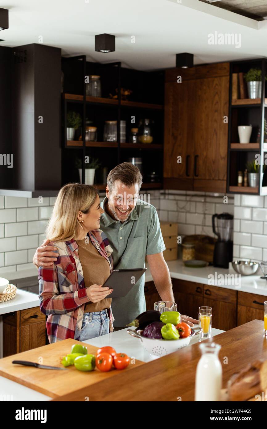 A cheerful couple stands in a well-lit kitchen, engrossed in a digital ...