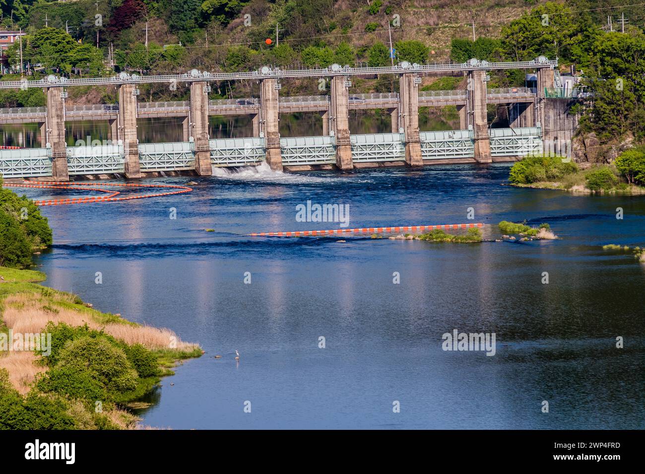 Landscape Of Dam Releasing Water Through One Floodgate Into River With Blue Water Located Near