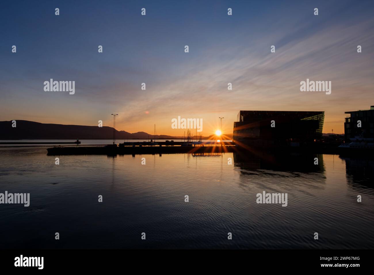 Sun rising behind mountains near Reykjavik casts rays over calm water ...