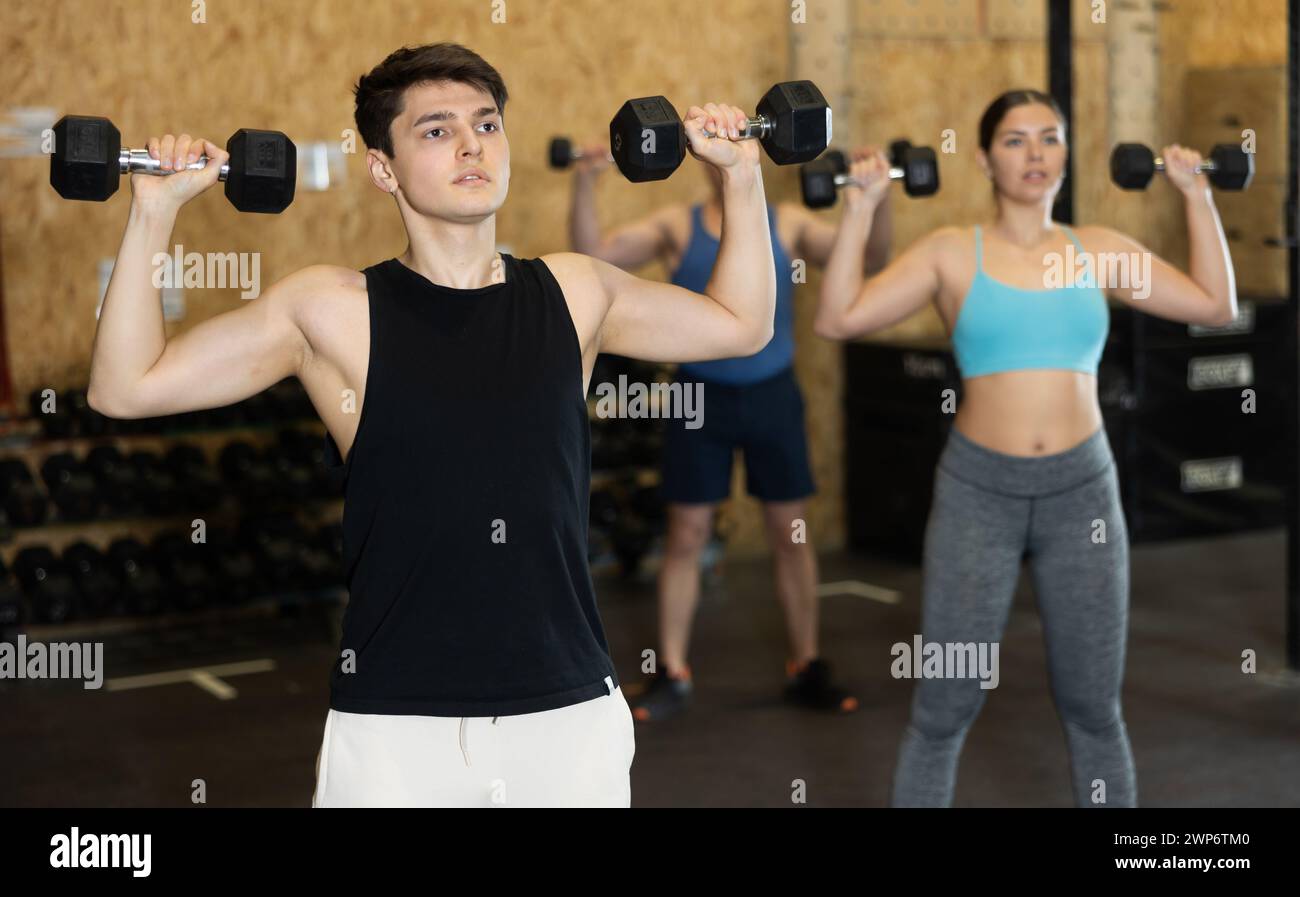 Motivated young man practicing exercises with dumbbells near other ...