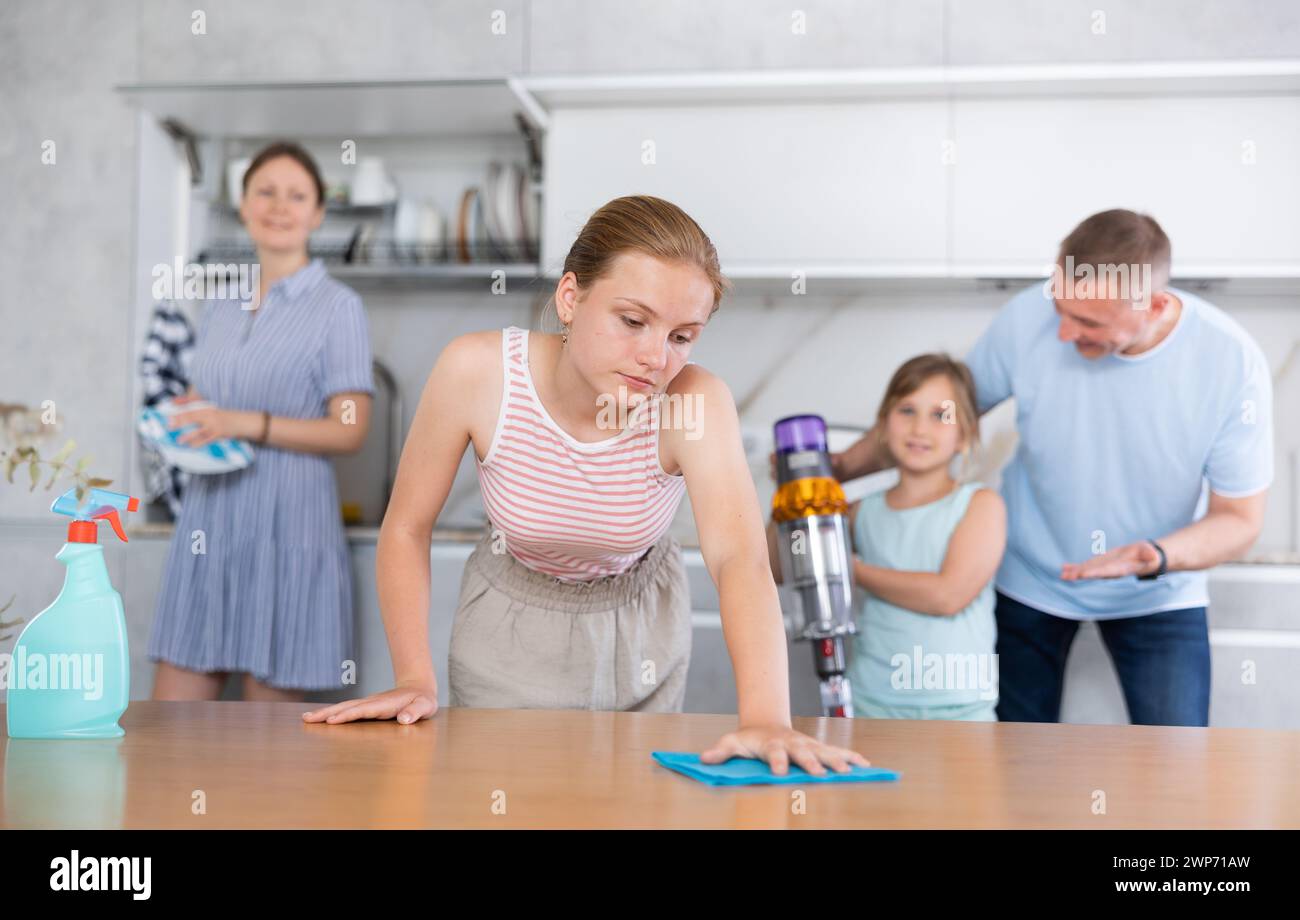 Teen girl cleans table with rag, family clean kitchen in background ...