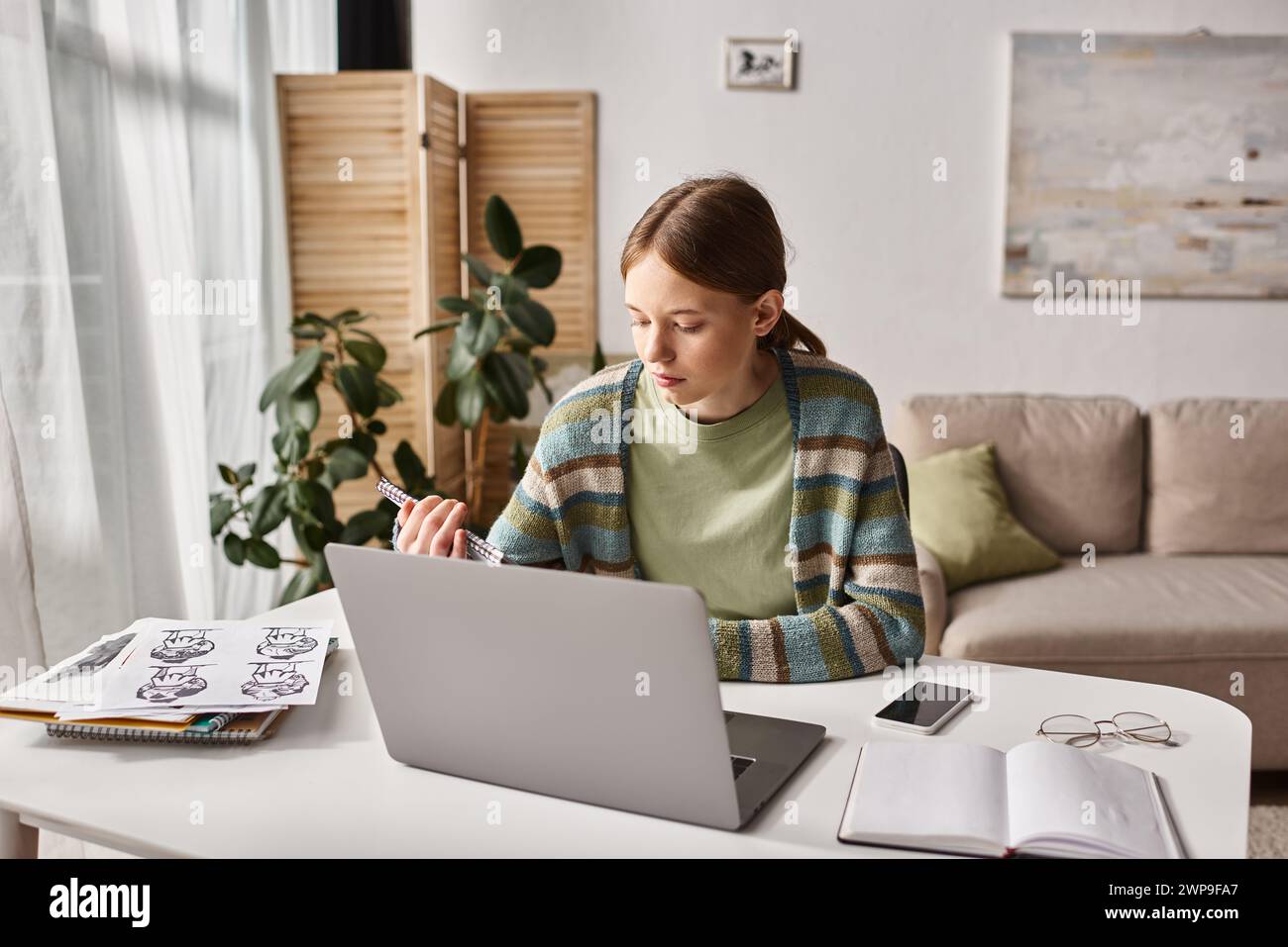 Focused teen girl e-learning with laptop near drawings and smartphone ...