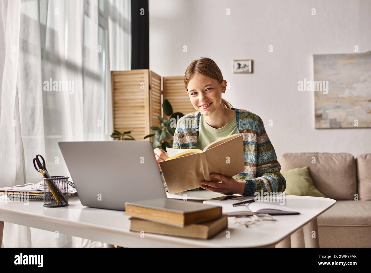 gen z girl with a book smiling while using a laptop for e-learning at ...