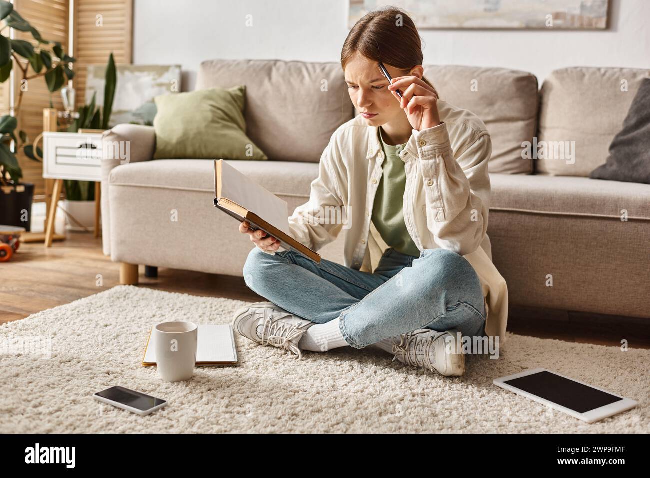 smart teen girl reading book while doing homework among devices and cup ...
