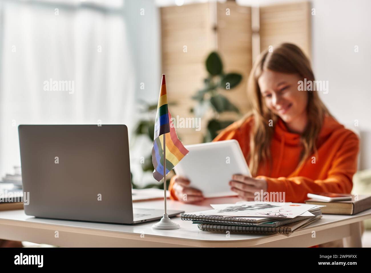 Cheerful teenage girl with tablet engaging in e-learning process near lgbtq pride flag on desk ...