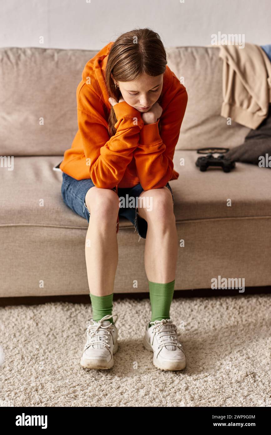 upset teenager girl in orange hoodie sitting on couch near blurred ...