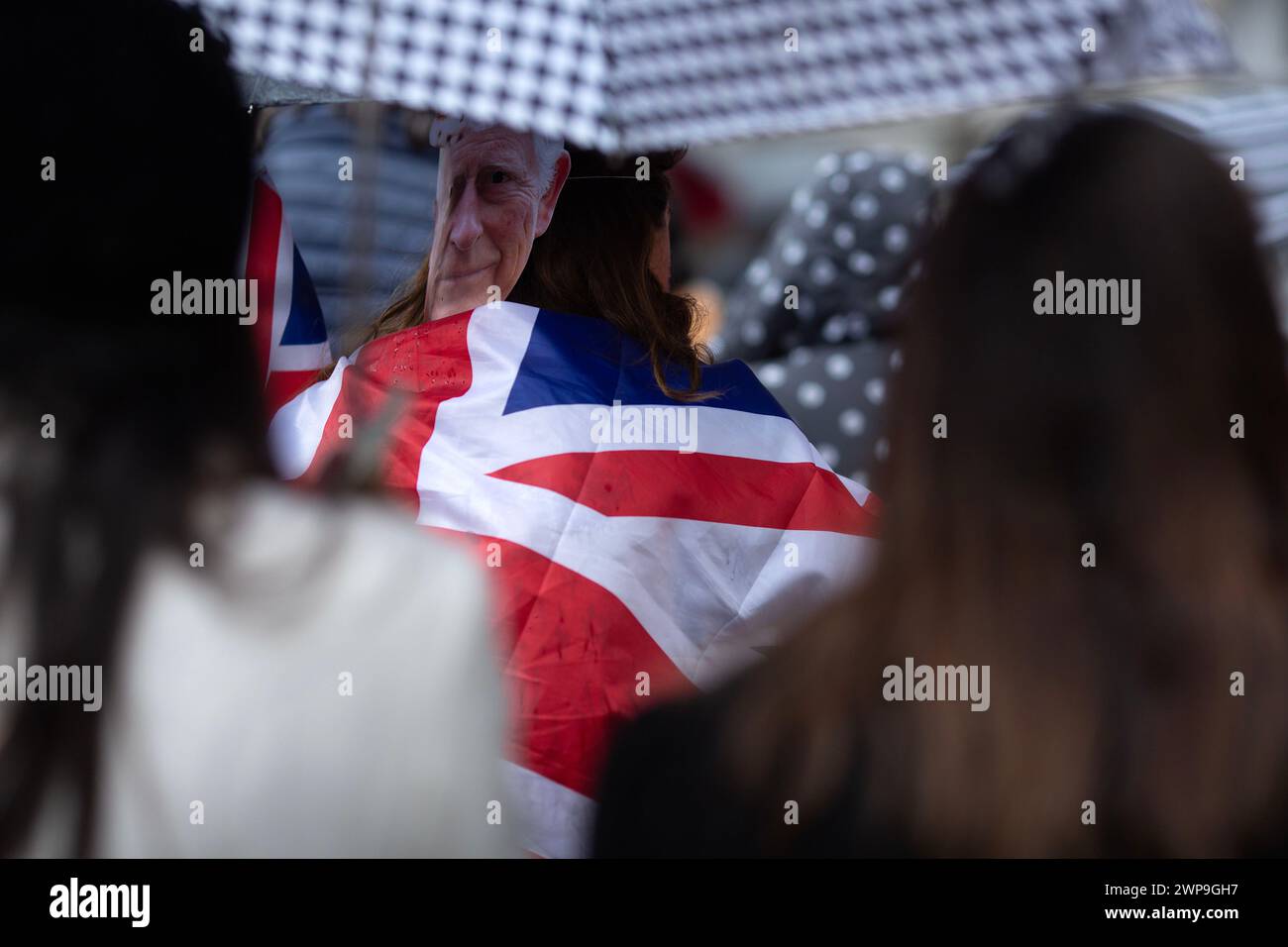 A mask of King Charles III is seen on the head of a pedestrian wearing ...