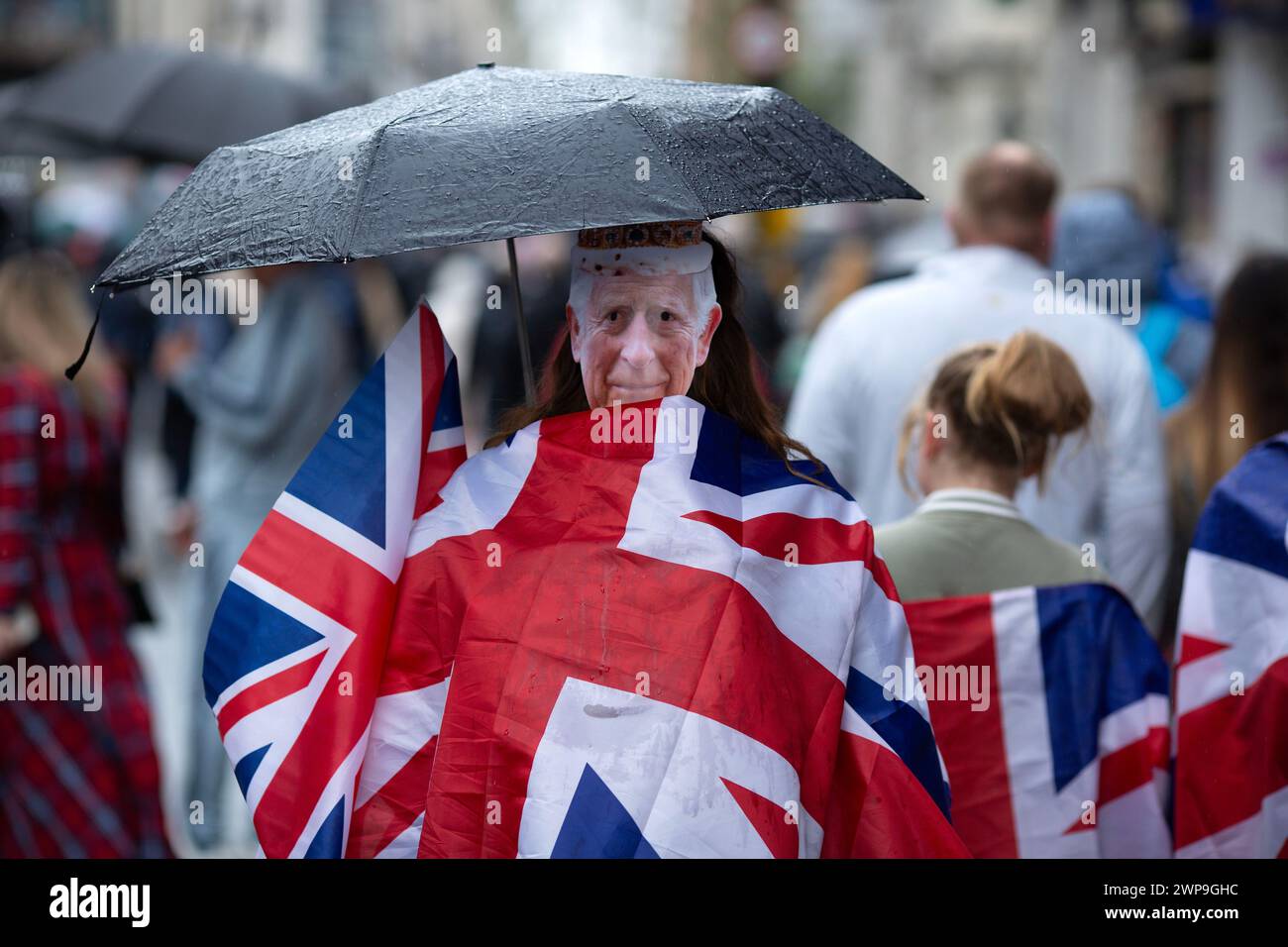 A mask of King Charles III is seen on the head of a pedestrian wearing ...