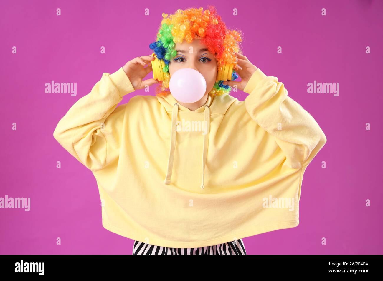 Trendy girl in clown costume blowing bubble gum while listening to ...