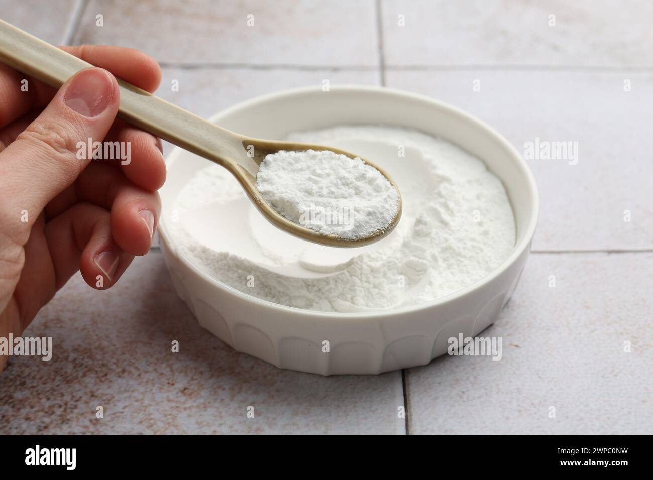 Woman taking baking powder with spoon from bowl at light tiled table ...