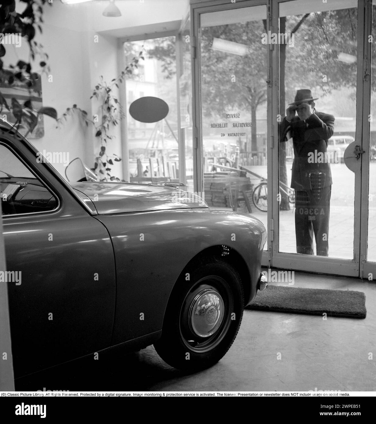 In the 1950s. A man is standing outside a store where an Alfa Romeo has ...