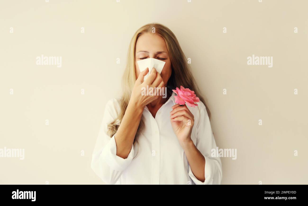 Sick exhausted woman suffering from pollen holds flower sneezing blow nose using tissue on white ...