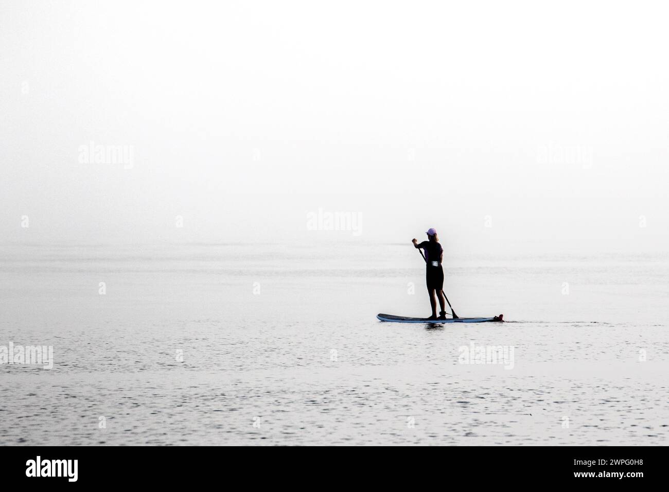 Silhouette of a peaceful solo woman on a paddleboard exploring alone in dense eerie fog on a ...