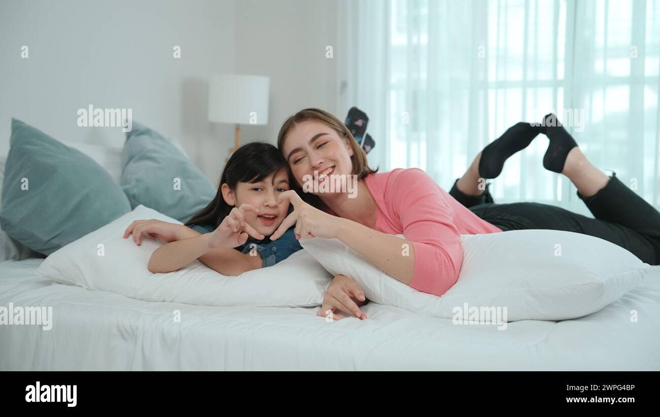Happy asian daughter and smart mom lie on bed while looking at camera ...