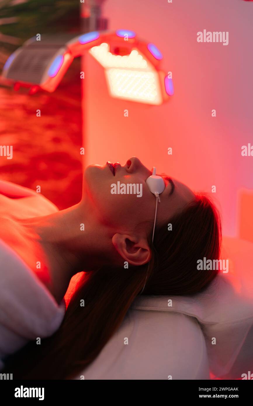 Vertical shot of young woman having red LED light facial photodynamic ...