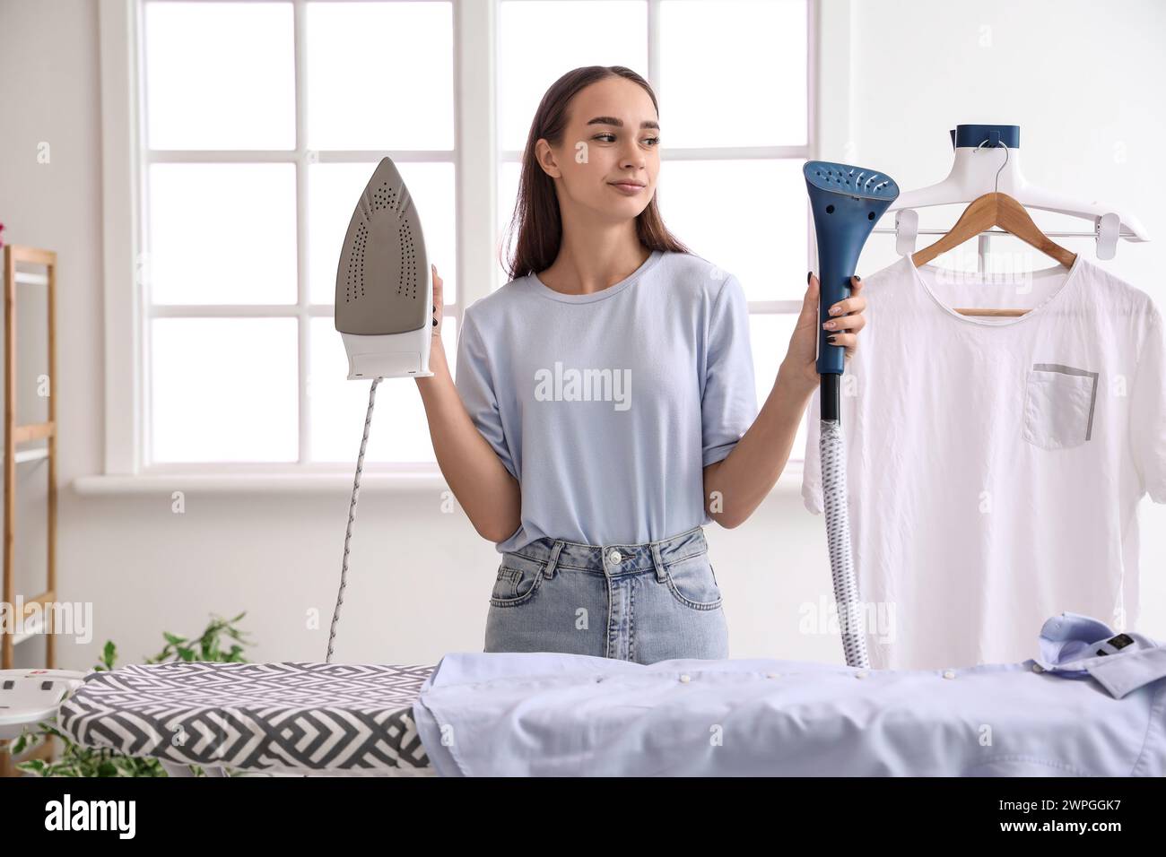 Pretty young woman with modern iron and garment steamer in laundry room ...