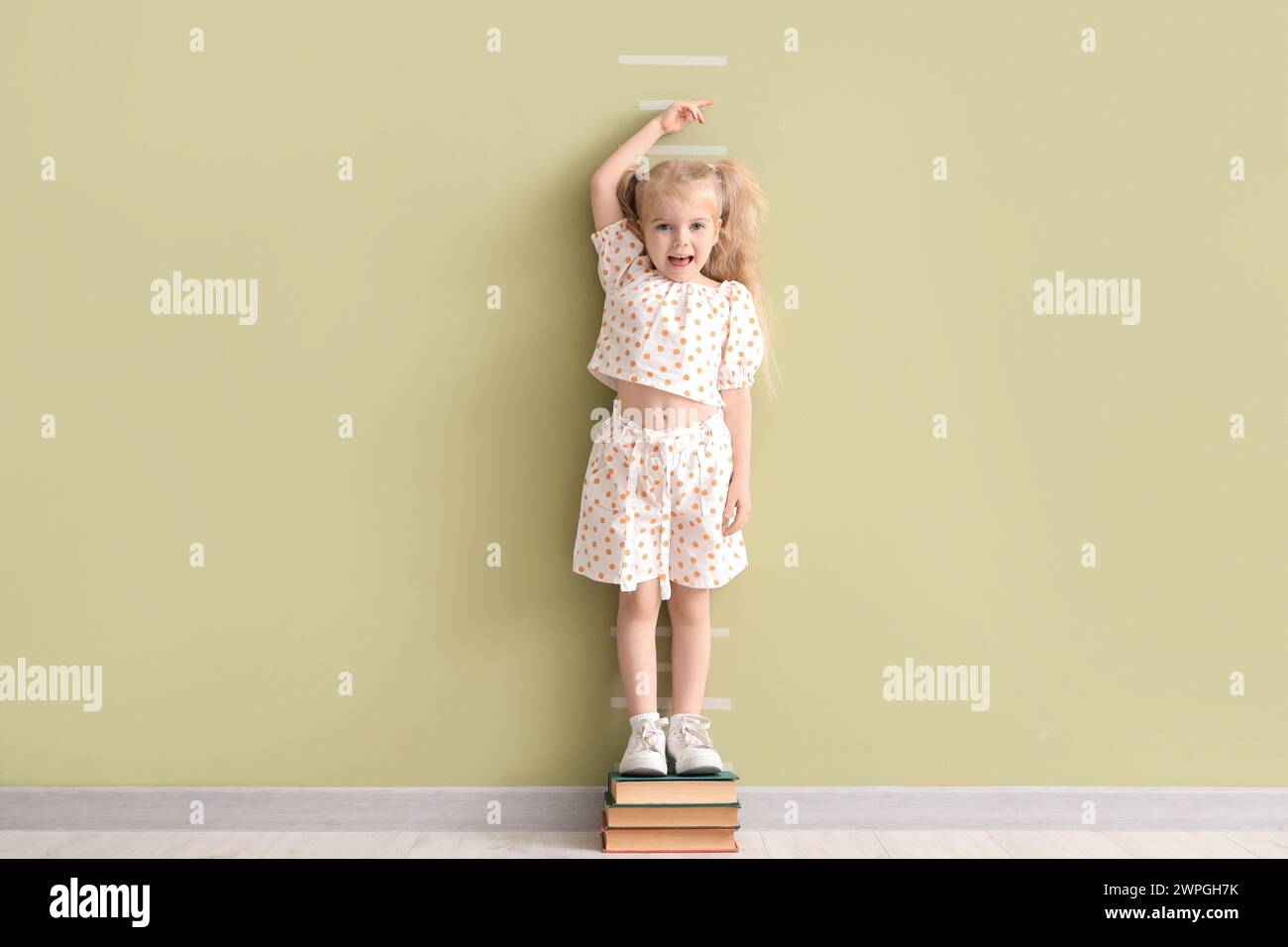 Cute little girl standing on books and measuring height near green wall ...