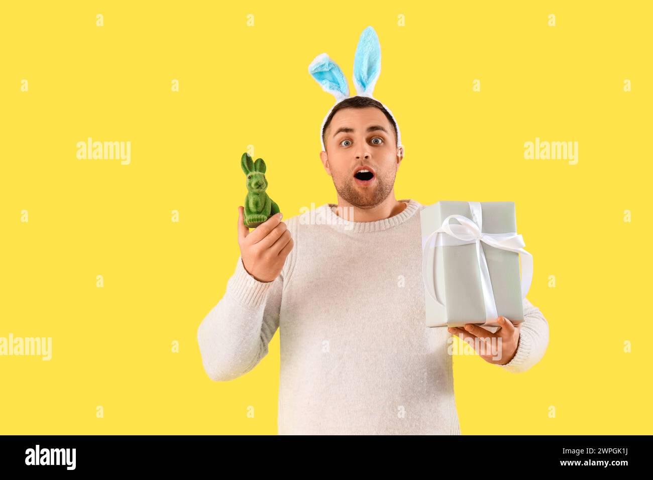 Shocked young man in bunny ears with gift box and toy rabbit on yellow ...
