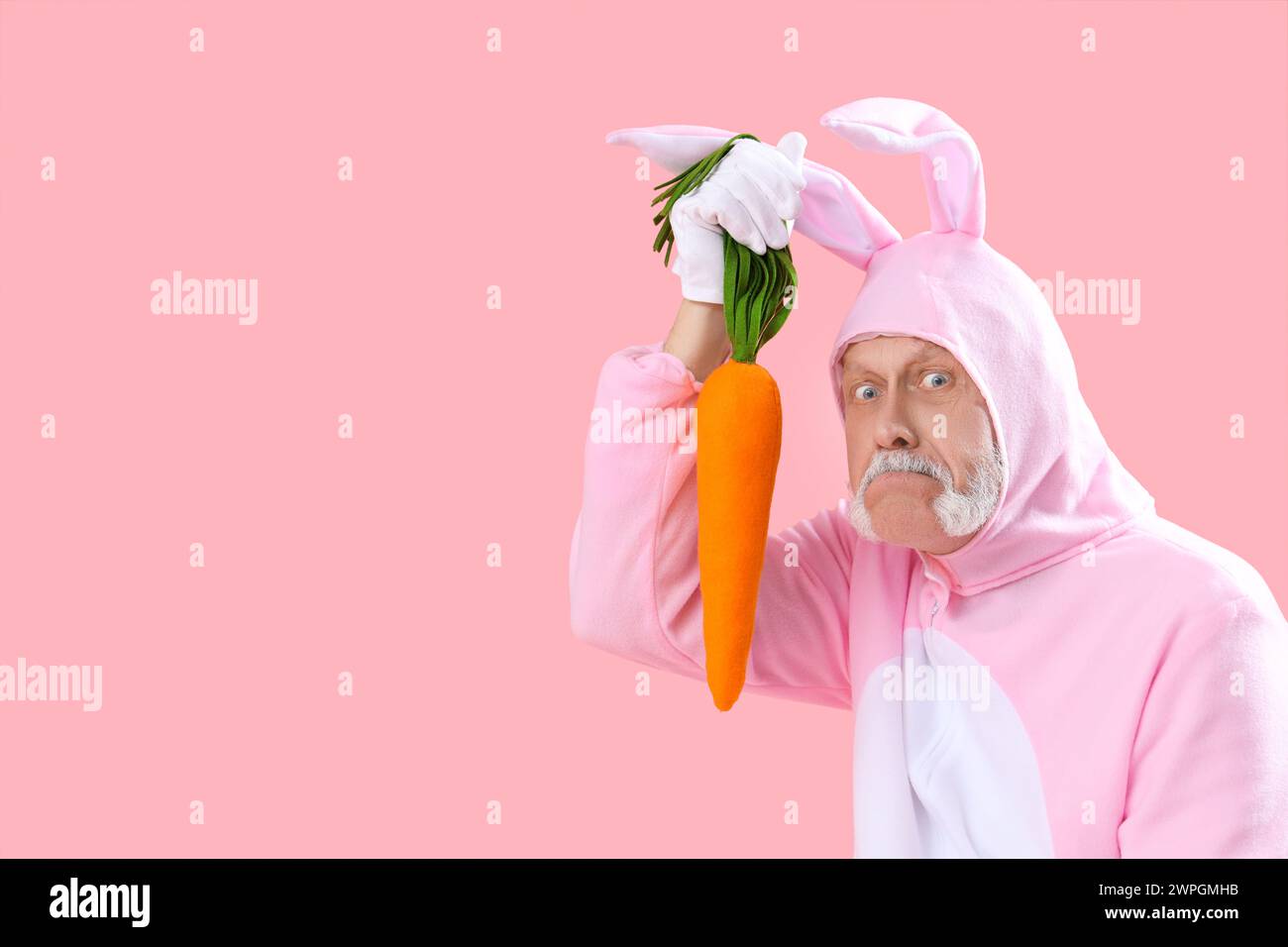 Senior man in Easter bunny costume with plush carrot on pink background ...