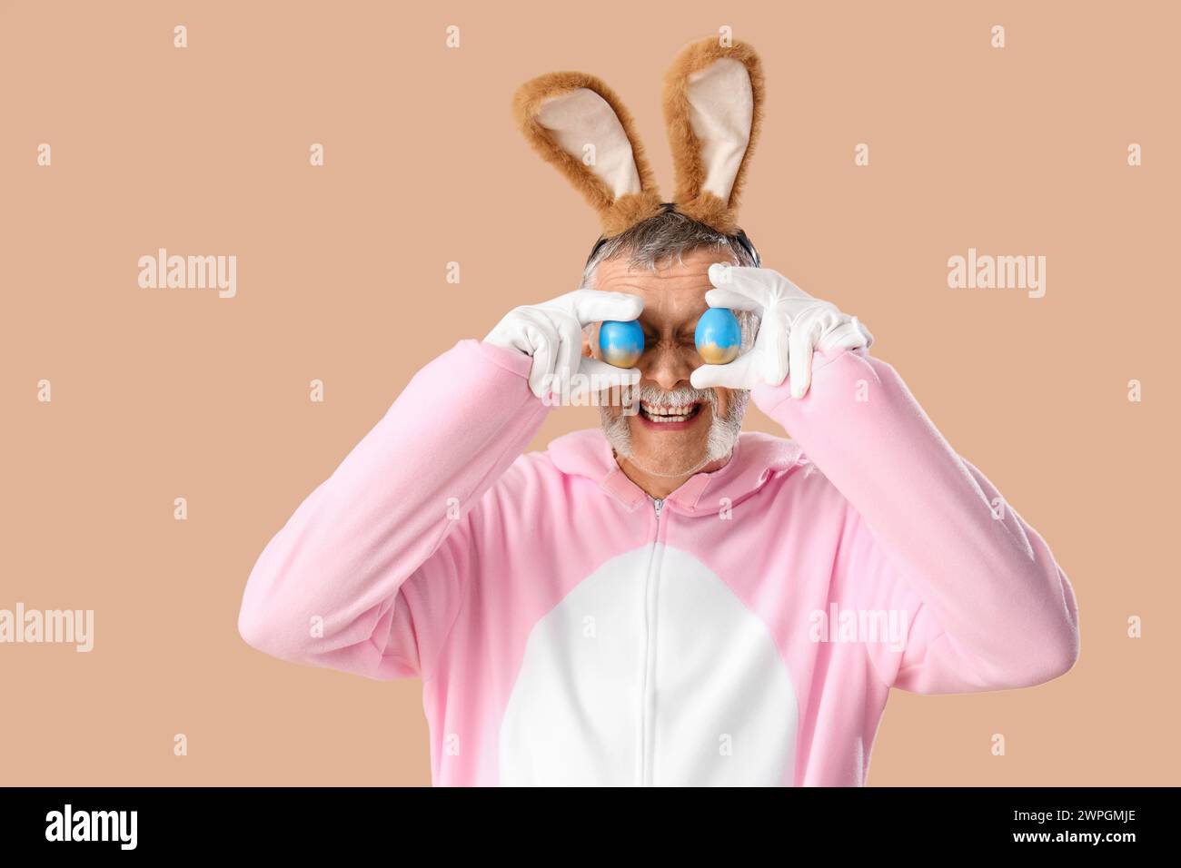 Senior man in bunny costume covering eyes with Easter eggs on beige ...