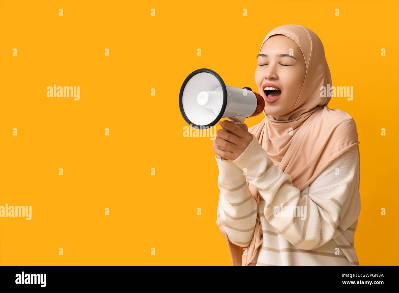 Young Asian Muslim woman shouting into megaphone on yellow background ...