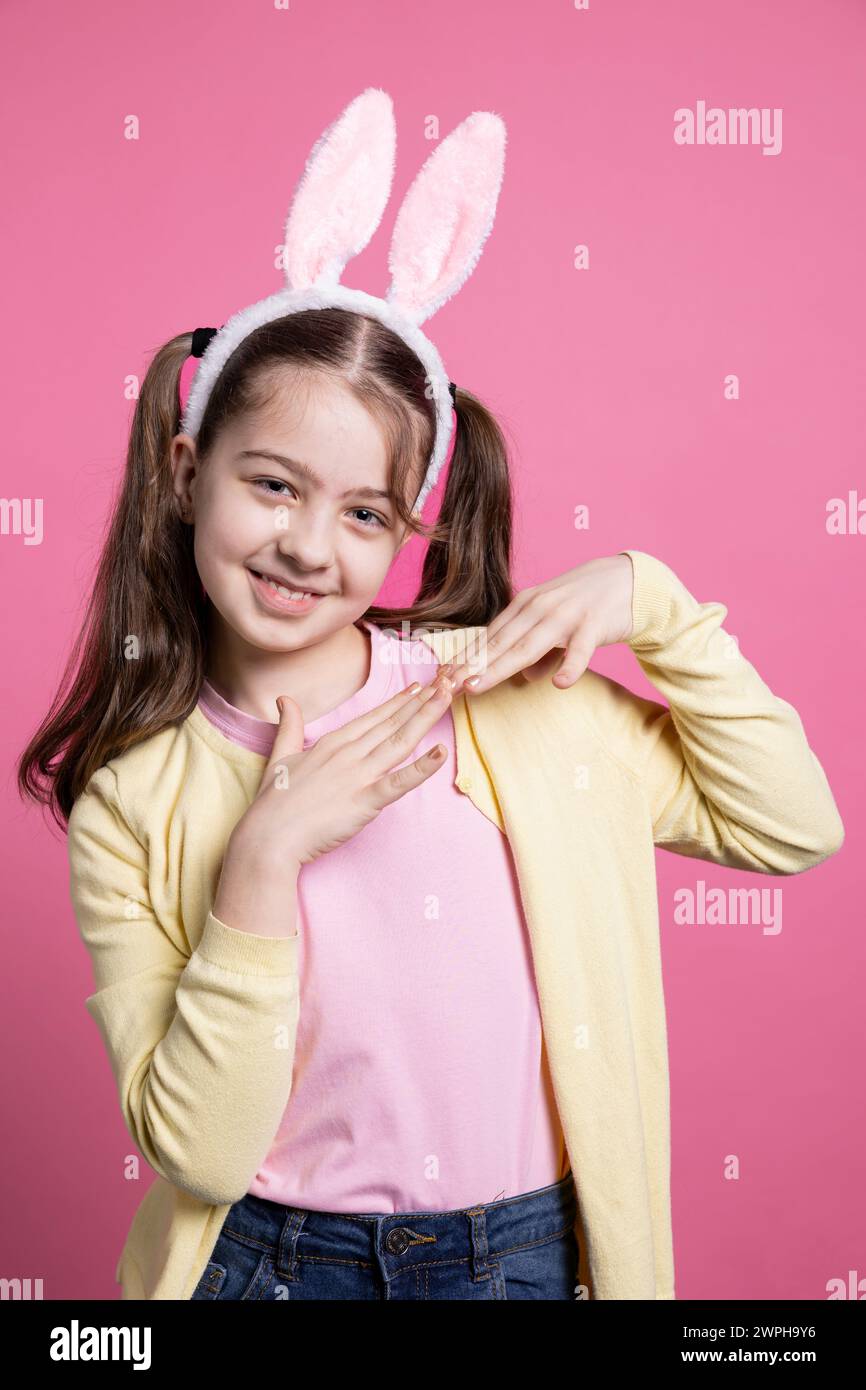 Cute young girl dancing and fooling around on camera, wearing bunny ears and pigtails over pink ...