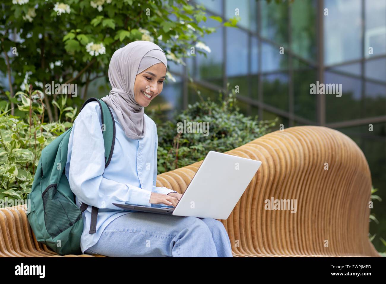 A cheerful Muslim woman wearing a hijab sits on a bench with a laptop ...