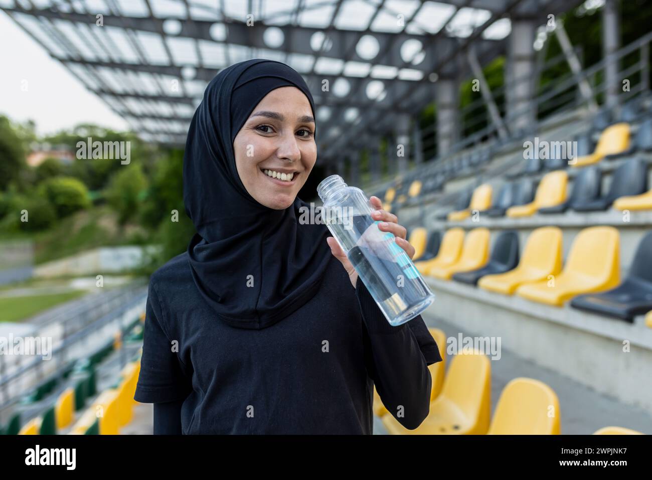 A happy Muslim woman in sports attire and hijab holding a water bottle ...