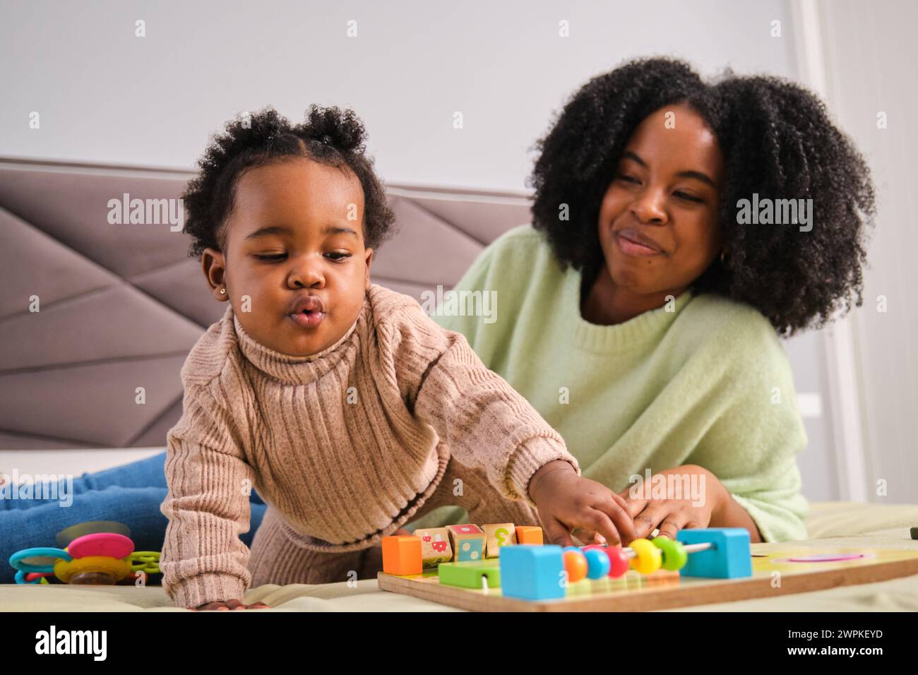 Cuban mother looking at her toddler daughter playing on bed at home ...