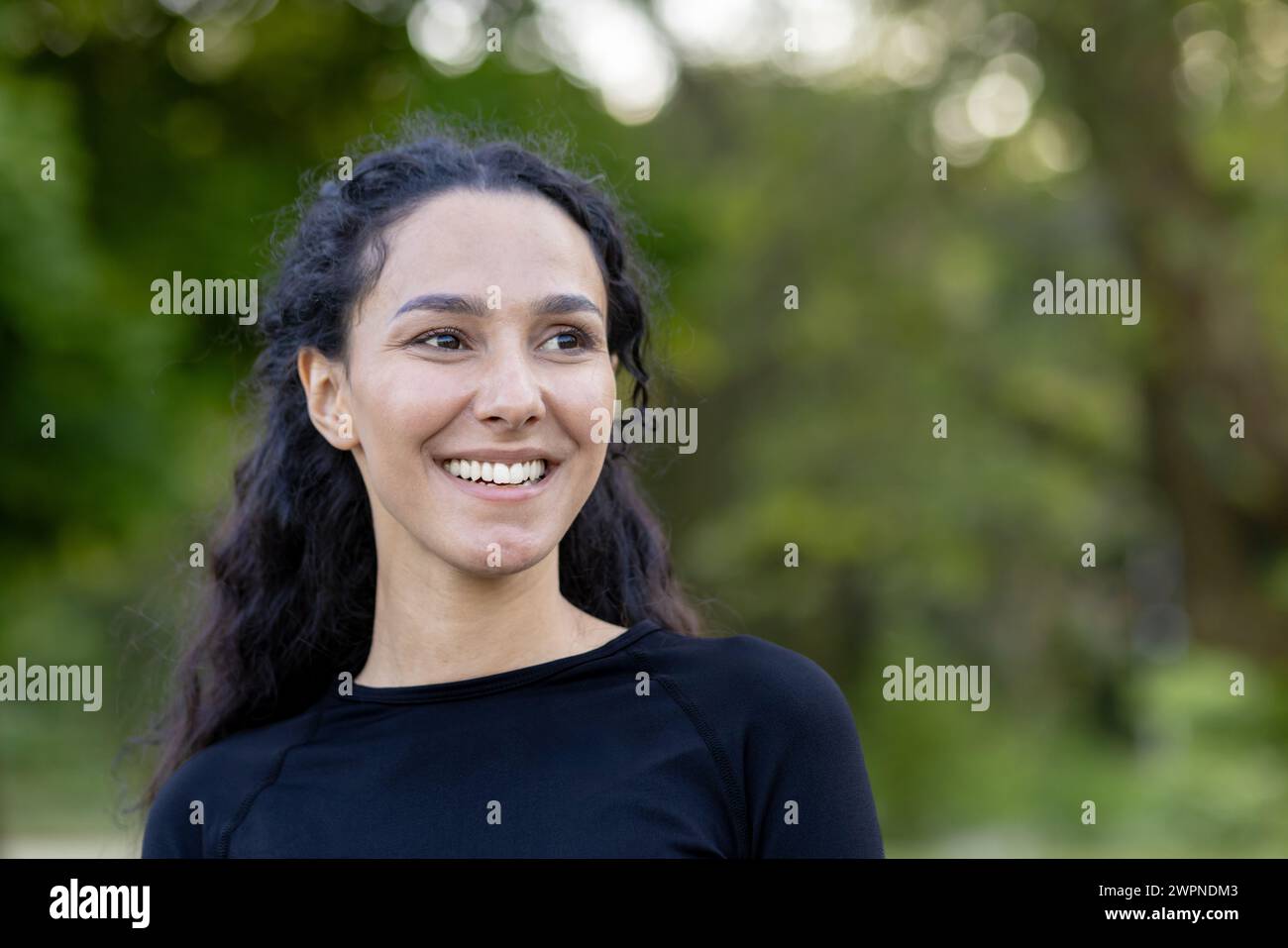 Portrait of a content young woman smiling gently, with green trees in ...