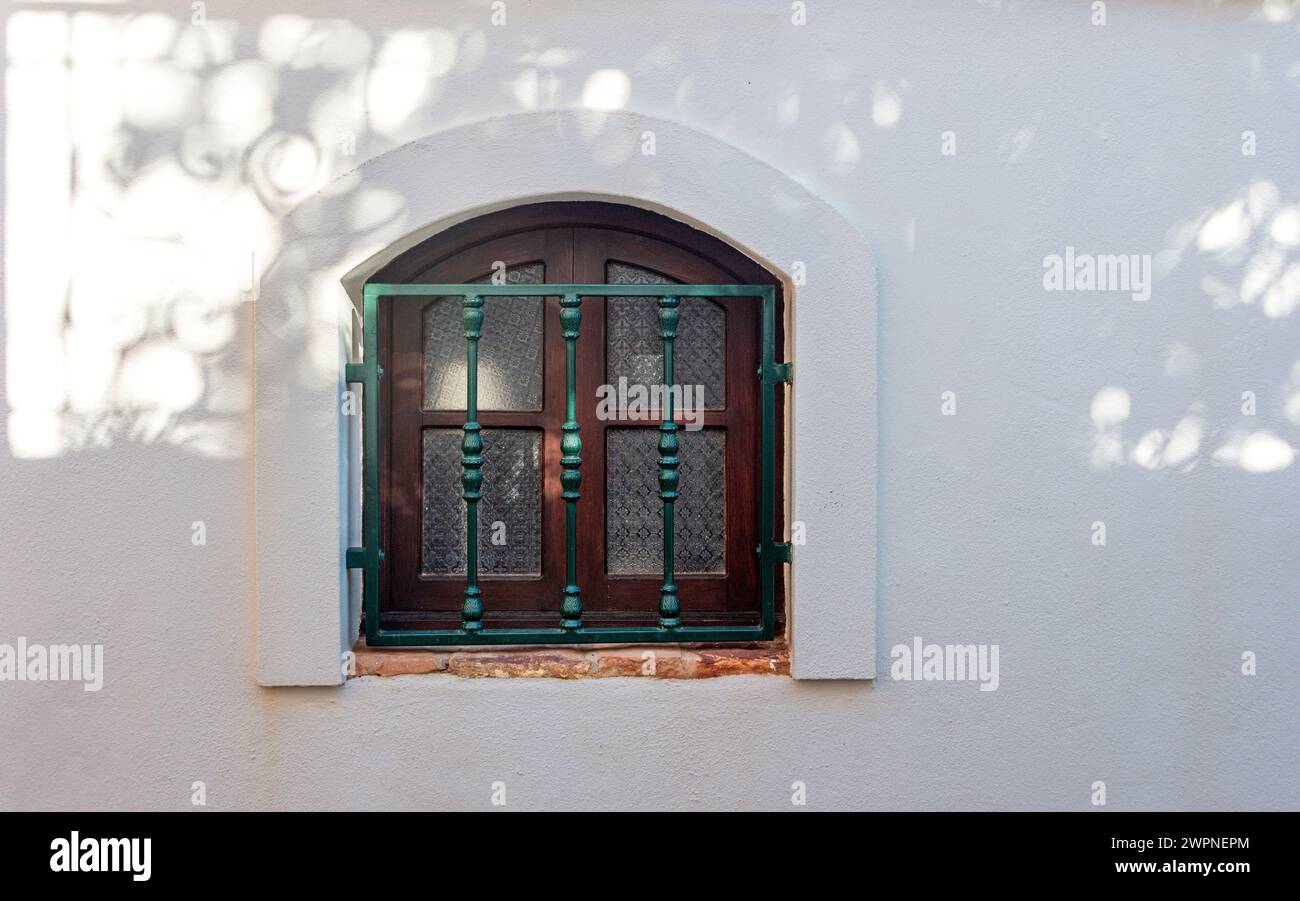 arched wooden window with grille set in a white wall in dappled ...